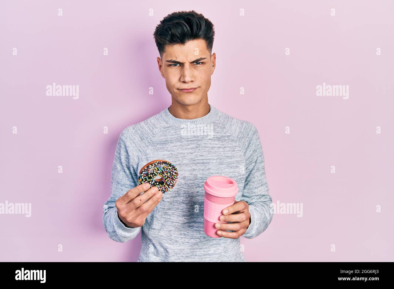 Young hispanic man eating doughnut and drinking coffee skeptic and ...