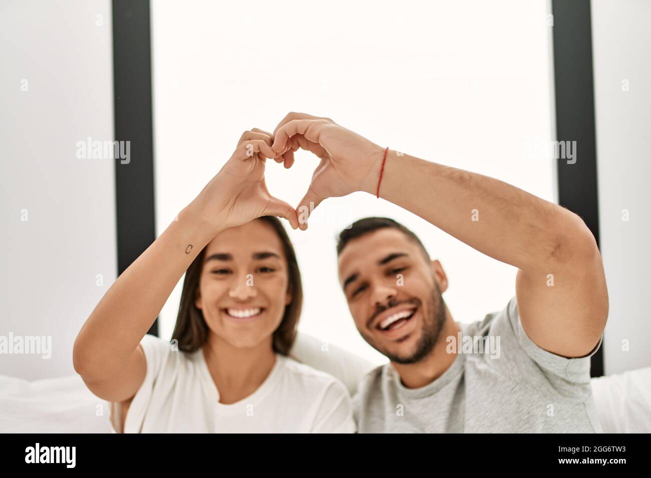 Young hispanic couple doing heart symbol with hands sitting on the bed ...