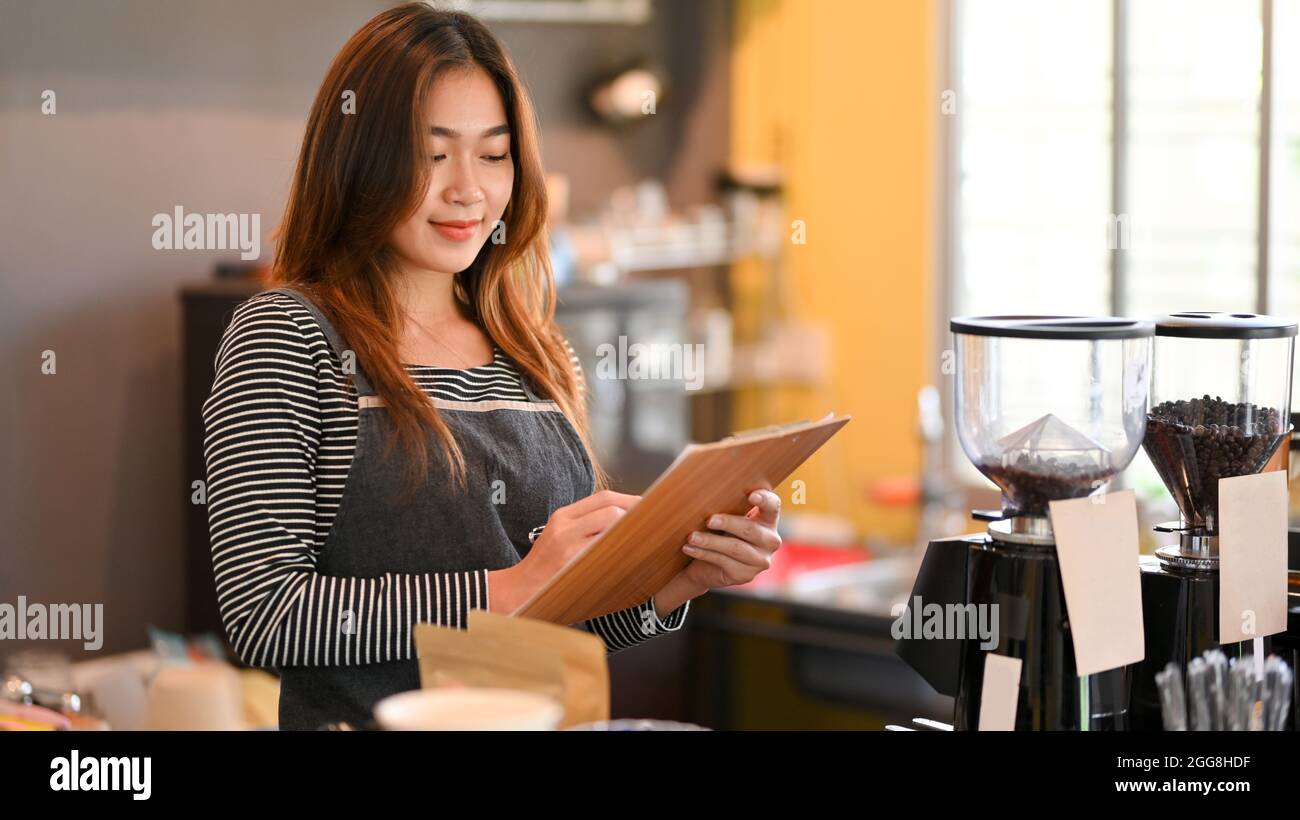 Pretty young barista looking at drinks menu, holding menu, taking order ...