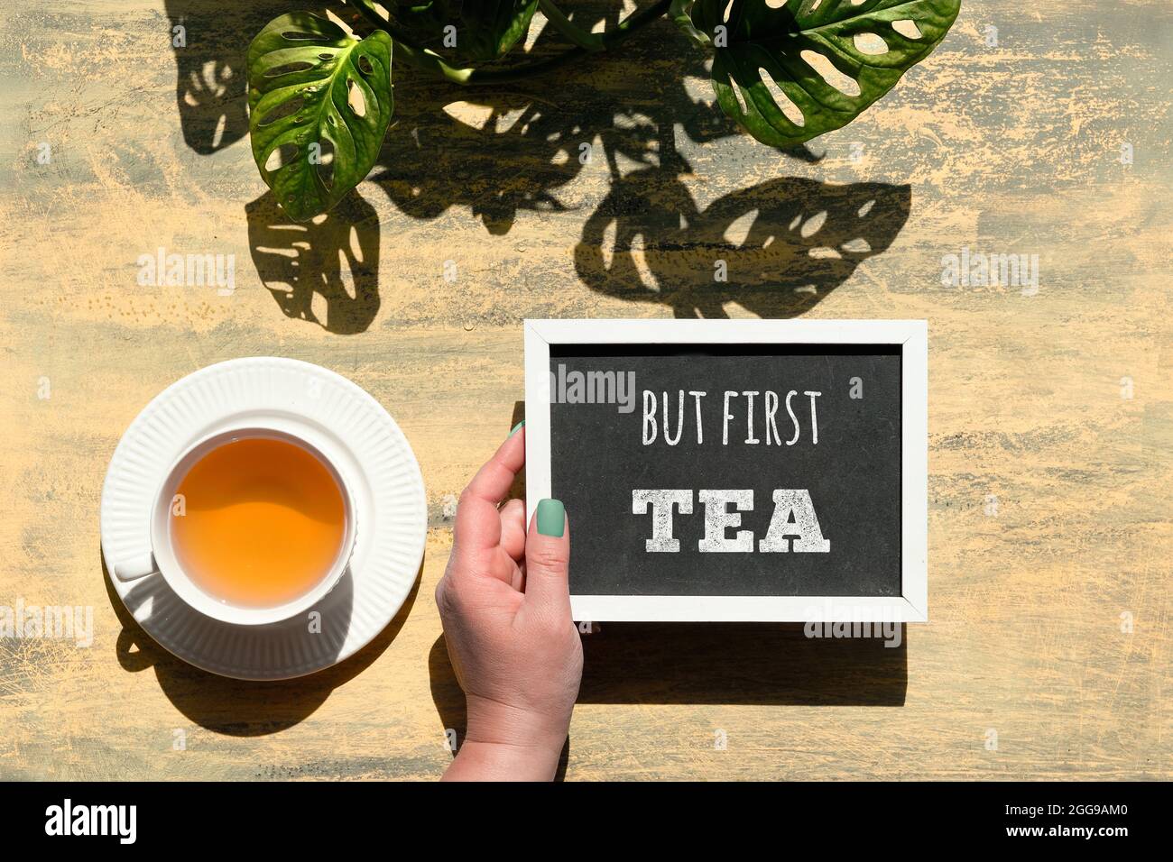 But first tea text on blackboard in hand. Cup of tea and monstera plant ...