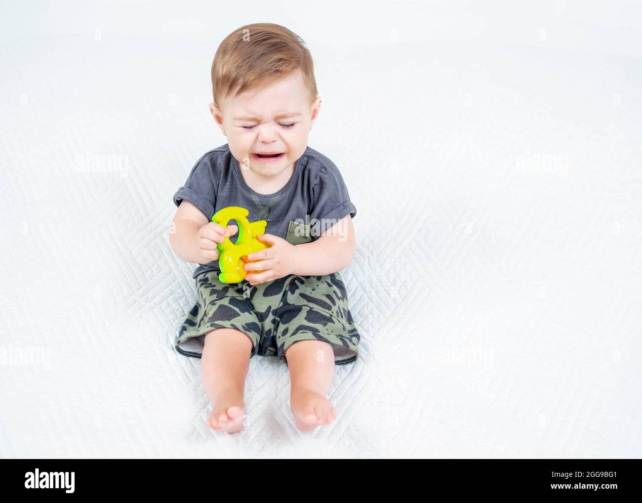 Sad baby boy in a khaki suit with colorful rattle toy sitting on bed ...