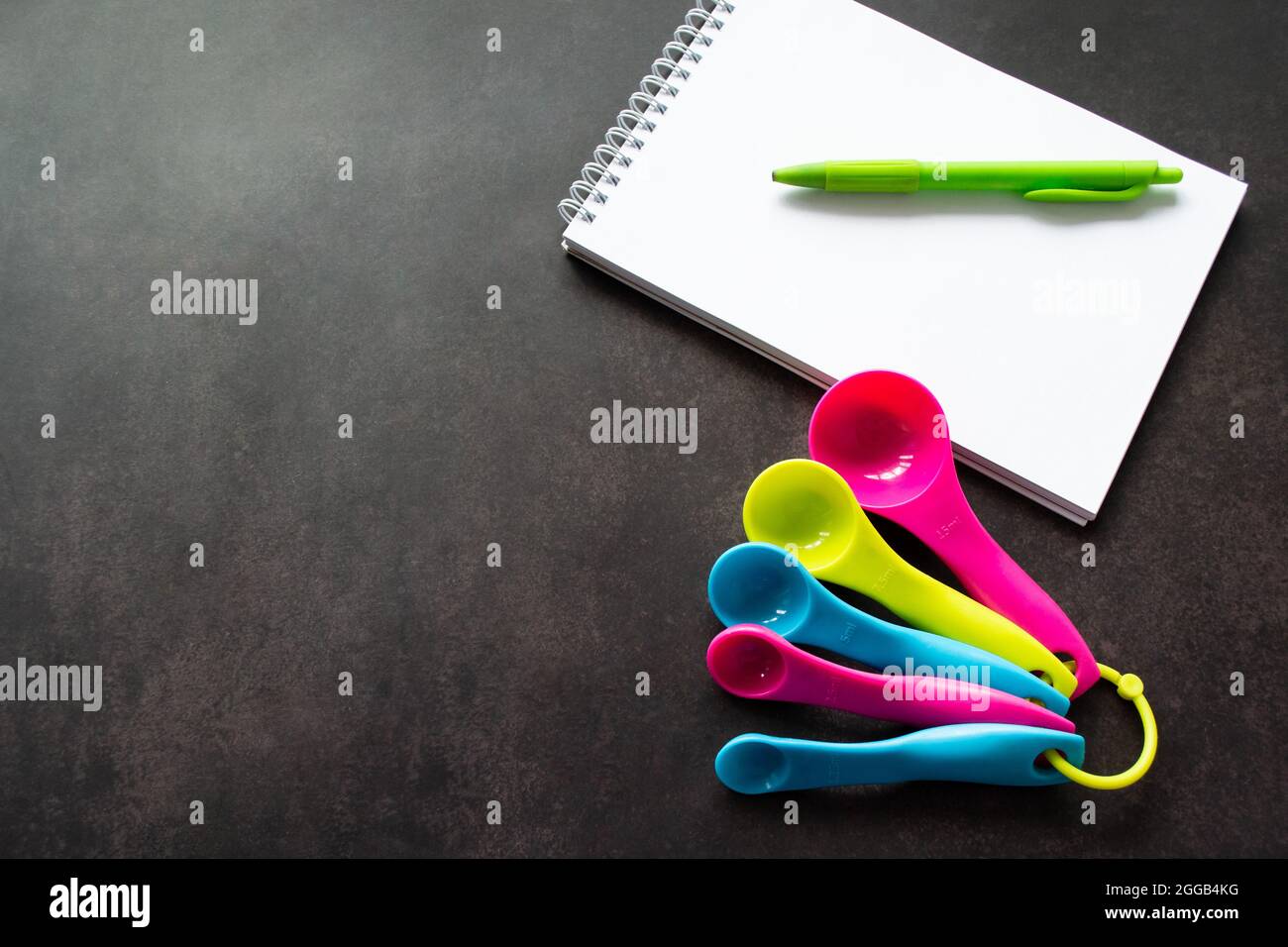 Multi colored measuring spoons, notebook with white pages and green pen ...
