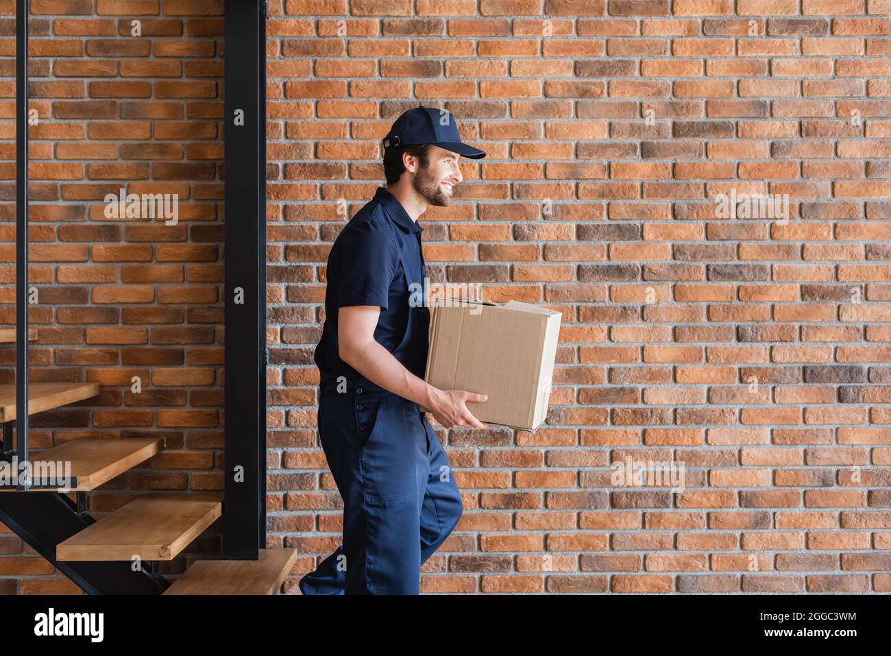 side view of smiling mover carrying carton box while walking downstairs ...