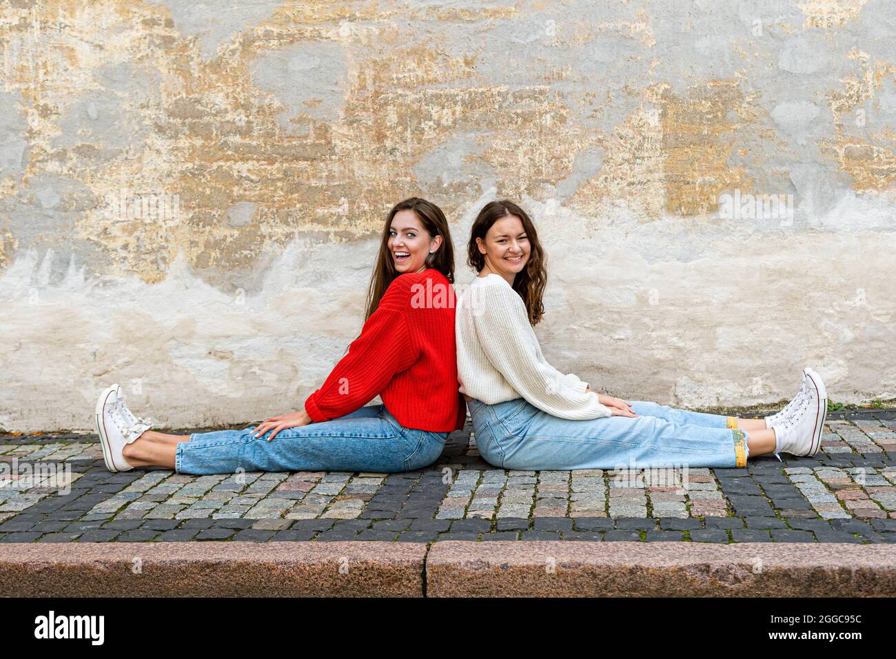 two young women sitting back to back on the pavement by an old wall ...
