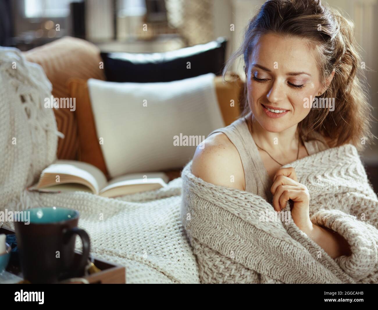 happy young woman in knitted cosy cardigan with mug sitting near sofa ...