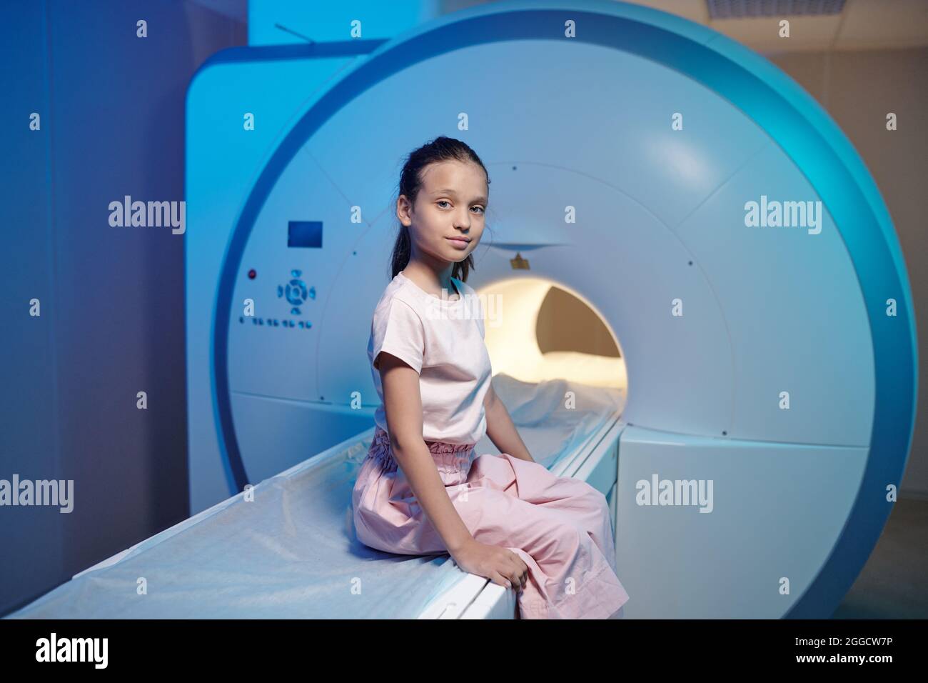 Cute little girl sitting on long table of mri scan machine in modern ...