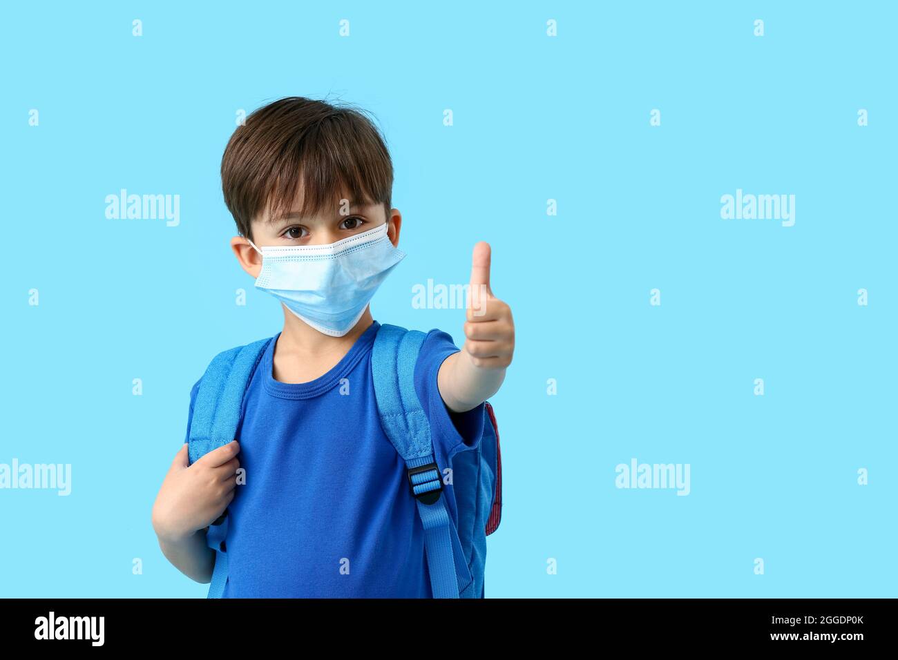Little schoolboy in medical mask showing thumb-up on color background ...