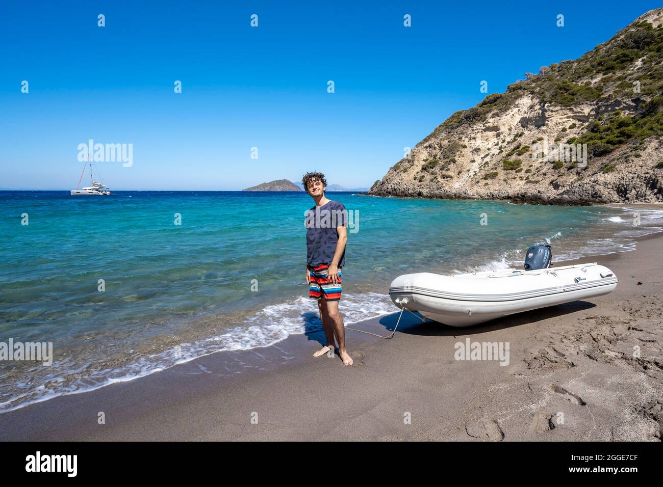 Young man with dinghy on a beach in Gyali, sailing boat behind ...