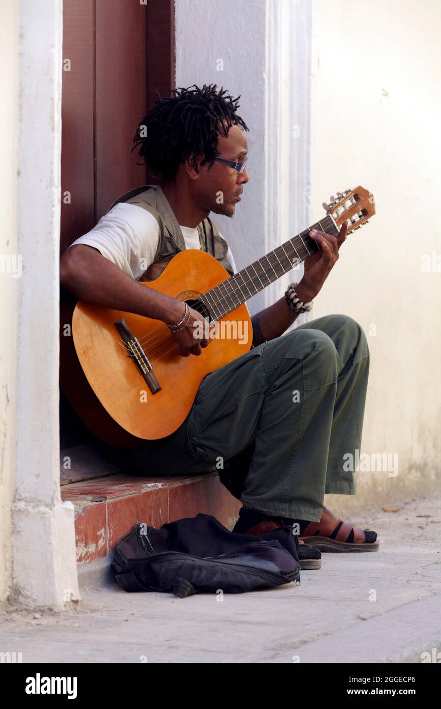 Young, dark-skinned man, Cuban, with rasta hairstyle sits in front door ...