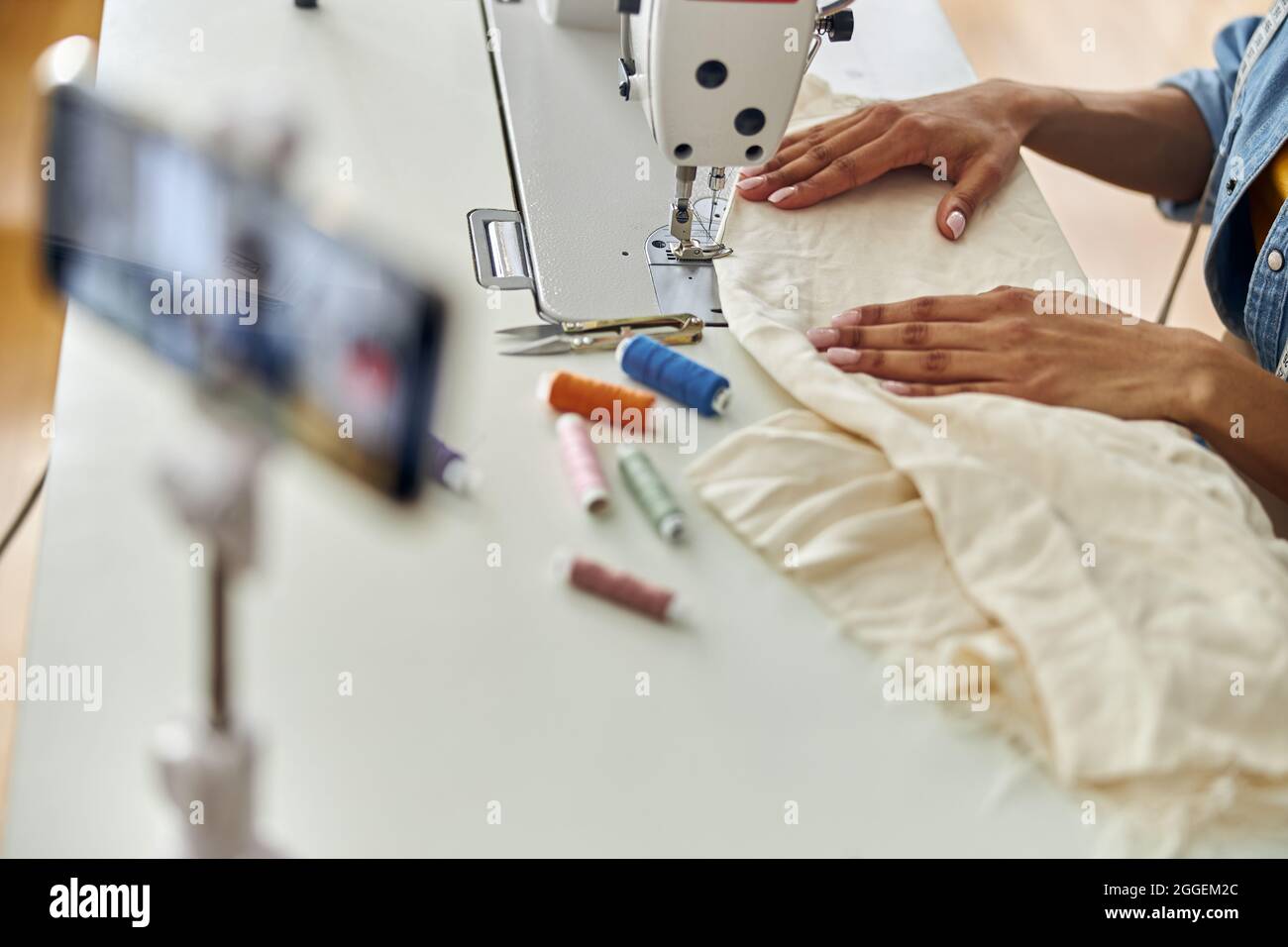 African-American dressmaker sews white fabric with new machine at table ...