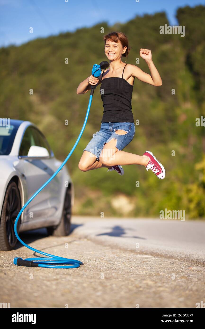 Beautiful young girl jumping next to an electric car. Holding blue ...