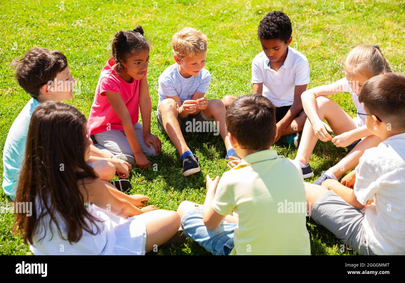 Group of happy kids resting on grass together and chatting in park ...