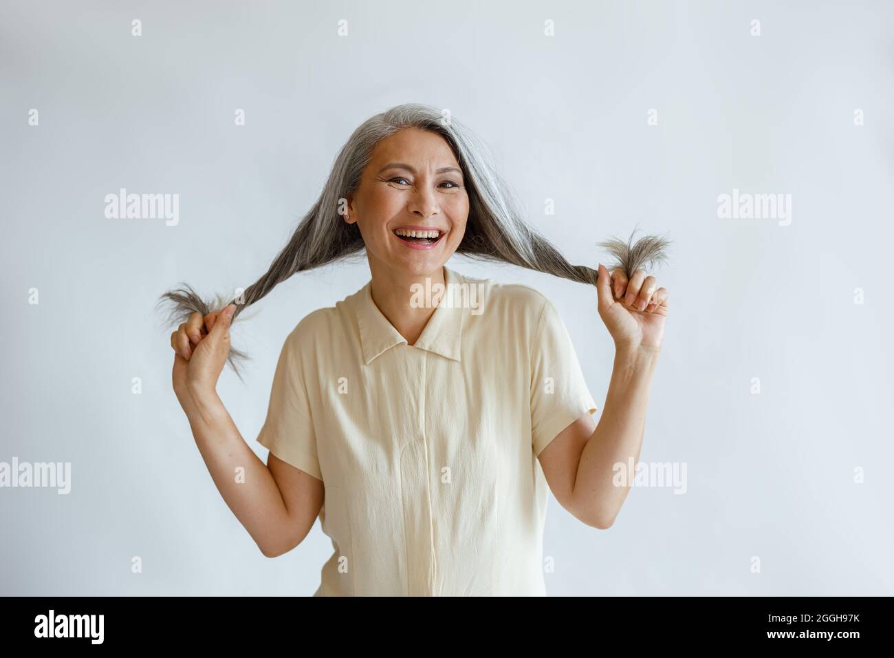 Happy middle aged Asian woman holds twisted hoary hair on light grey ...