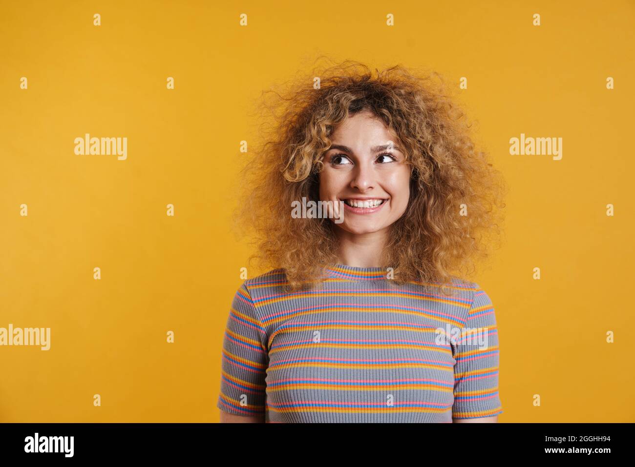 Happy smiling young blonde woman with fizzy hair in casual clothes standing over yellow wall ...