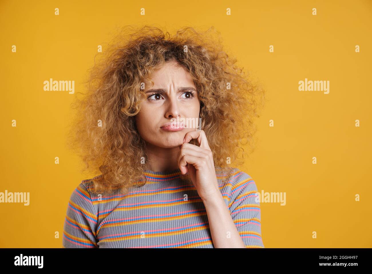 Pensive young casual woman with fizzy hairstyle standing over yellow wall background, looking ...