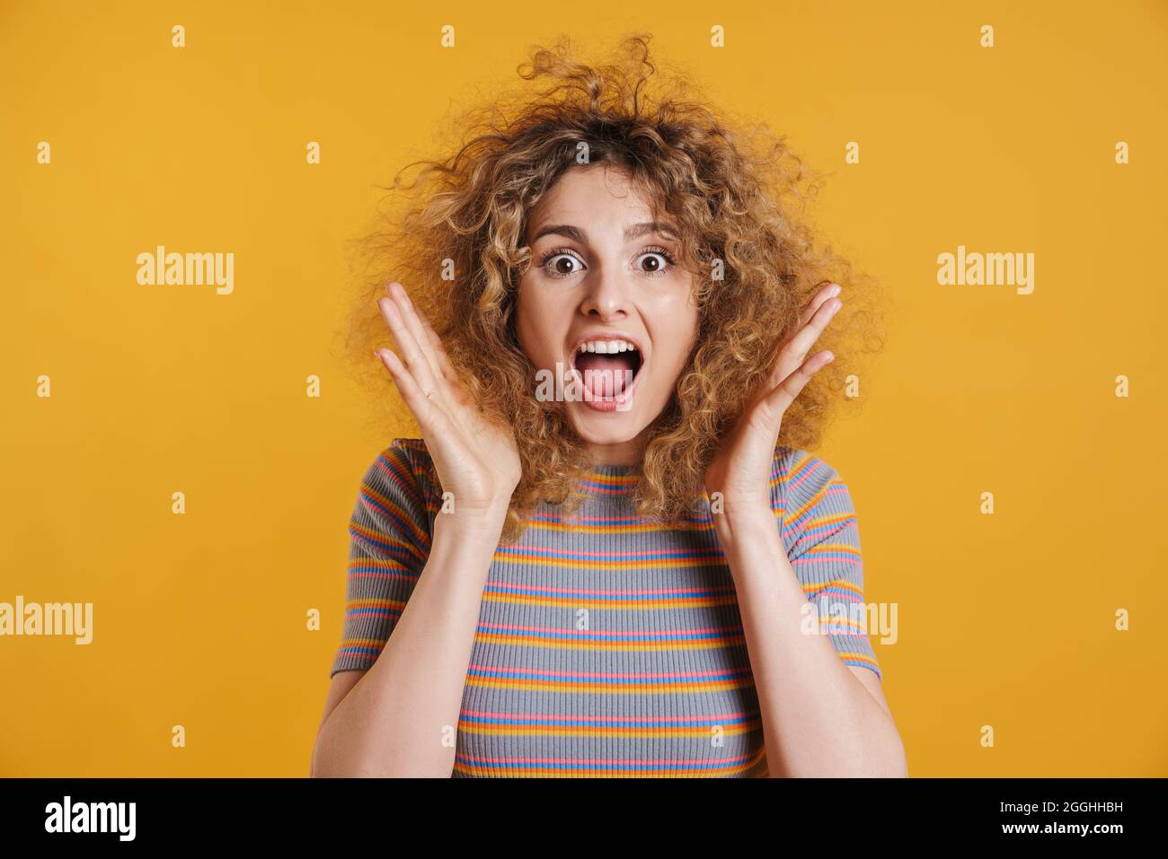 Happy excited young casual woman with fizzy hairstyle standing over yellow wall background ...