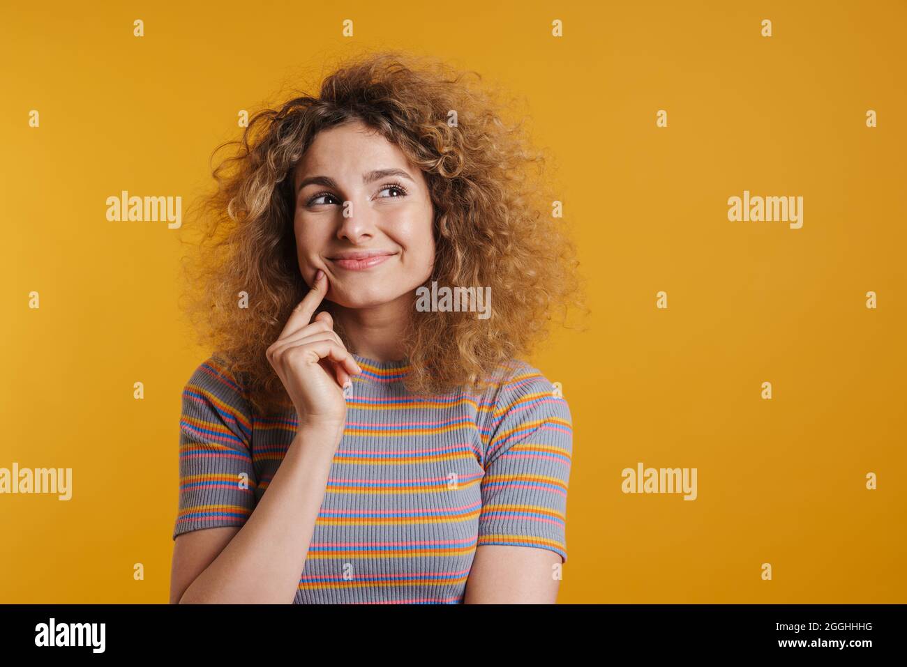 Pensive smiling young blonde woman with fizzy hair in casual clothes standing over yellow wall ...