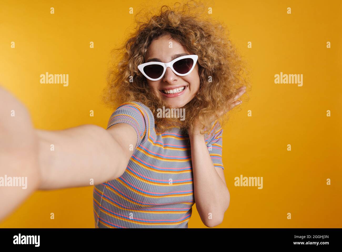 Happy young casual woman with fizzy hairstyle wearing sunglasses standing over yellow wall ...