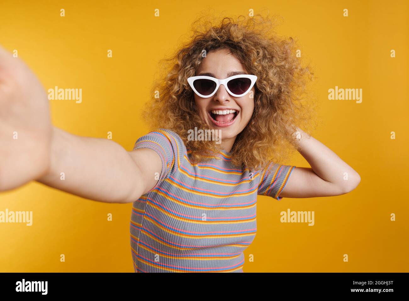 Happy young casual woman with fizzy hairstyle wearing sunglasses standing over yellow wall ...