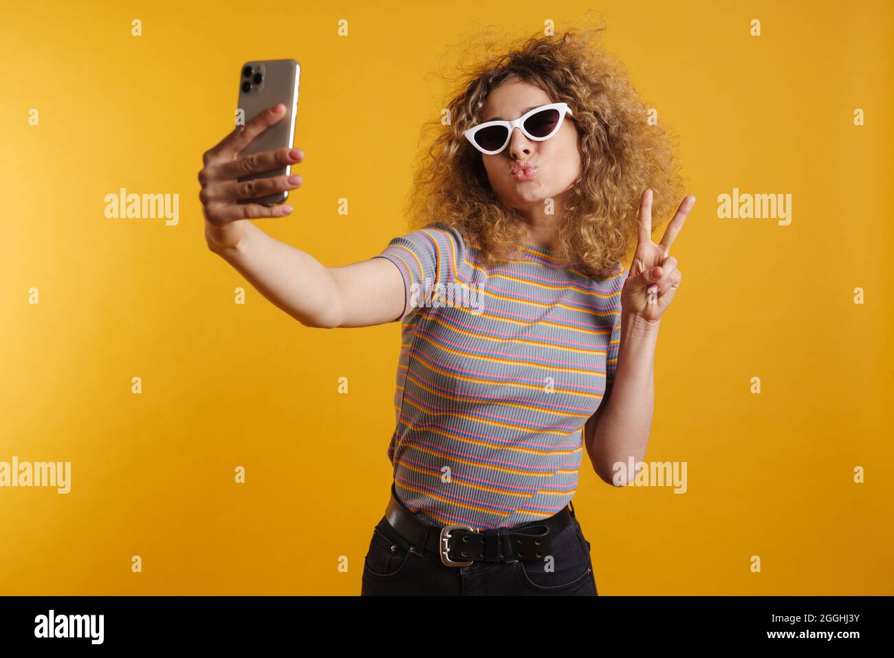 Young casual woman with fizzy hairstyle wearing sunglasses standing over yellow wall background ...