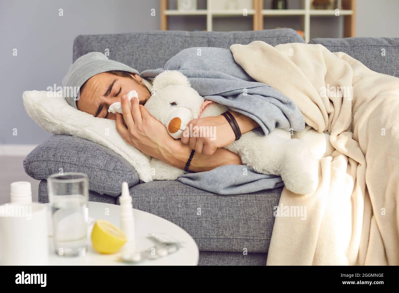 Sick young man lying on couch at home, hugging teddy and blowing nose ...