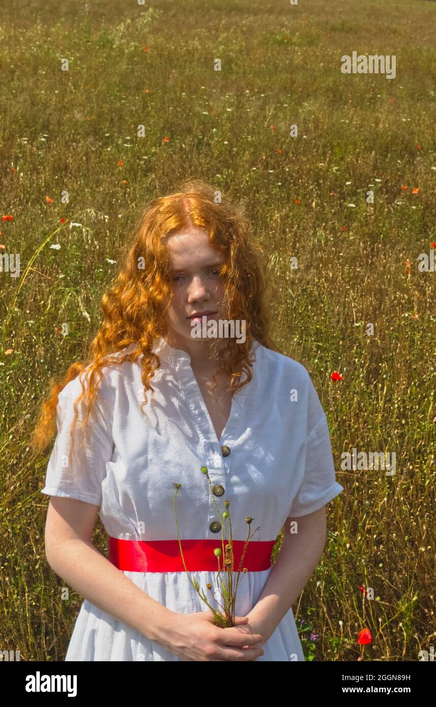 red haired girl wearing white dress in a field of dead poppies holding ...