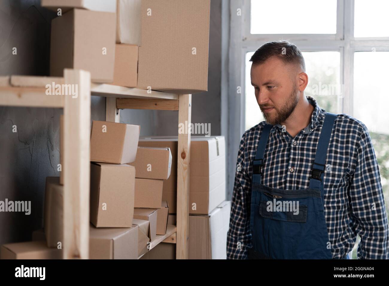 Young man dressed in blue overalls near cardboard boxes in a warehouse ...