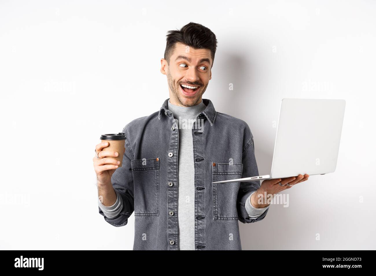 Cheerful young man working on laptop and drinking coffee, looking happy ...