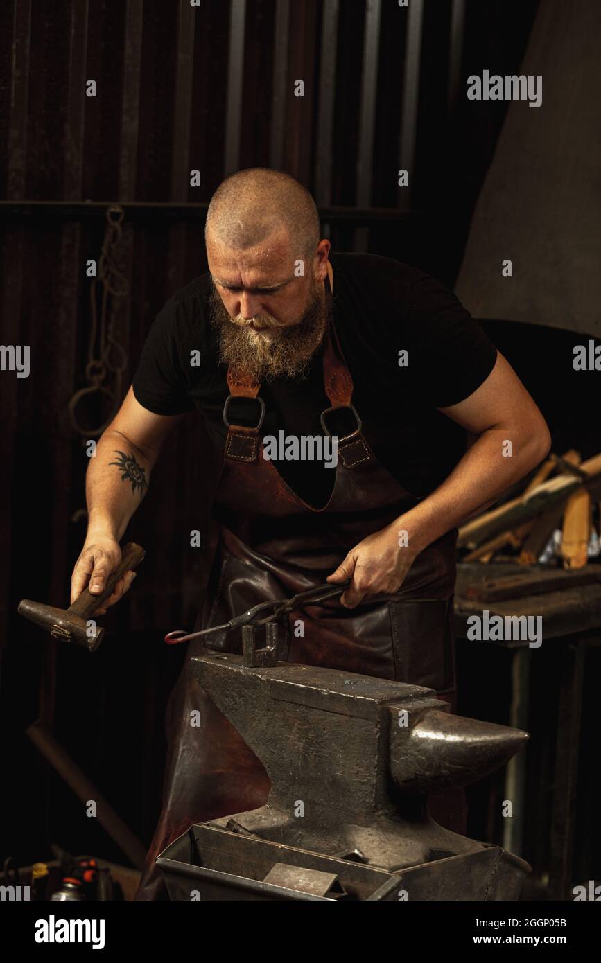 Bearded man, blacksmith manually forging the molten metal on the anvil ...