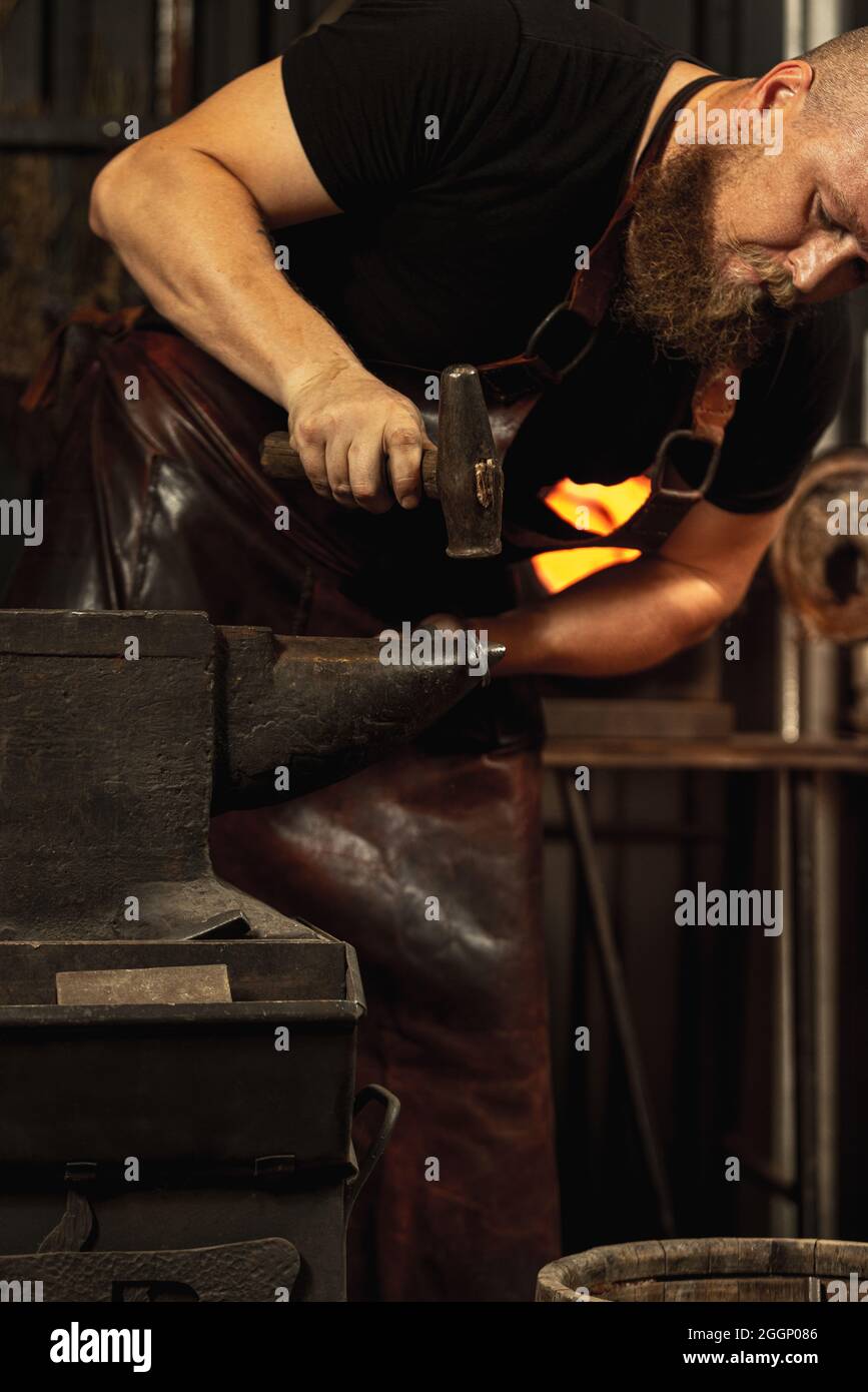 Bearded man, blacksmith manually forging the molten metal on the anvil ...