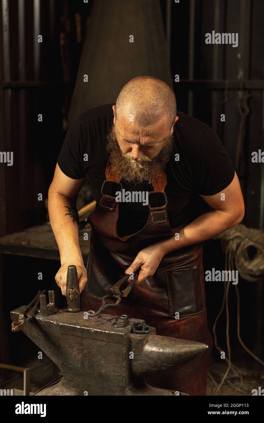 Bearded man, blacksmith manually forging the molten metal on the anvil ...