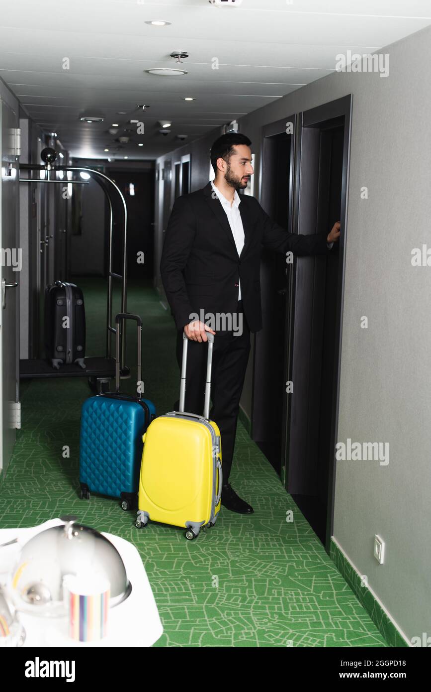 full length of bellboy in suit standing near luggage and knocking door ...