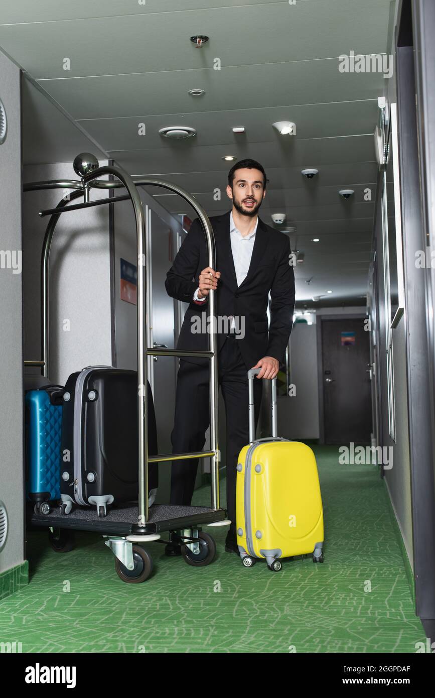 full length of bearded bellboy near metallic bell cart with luggage in ...