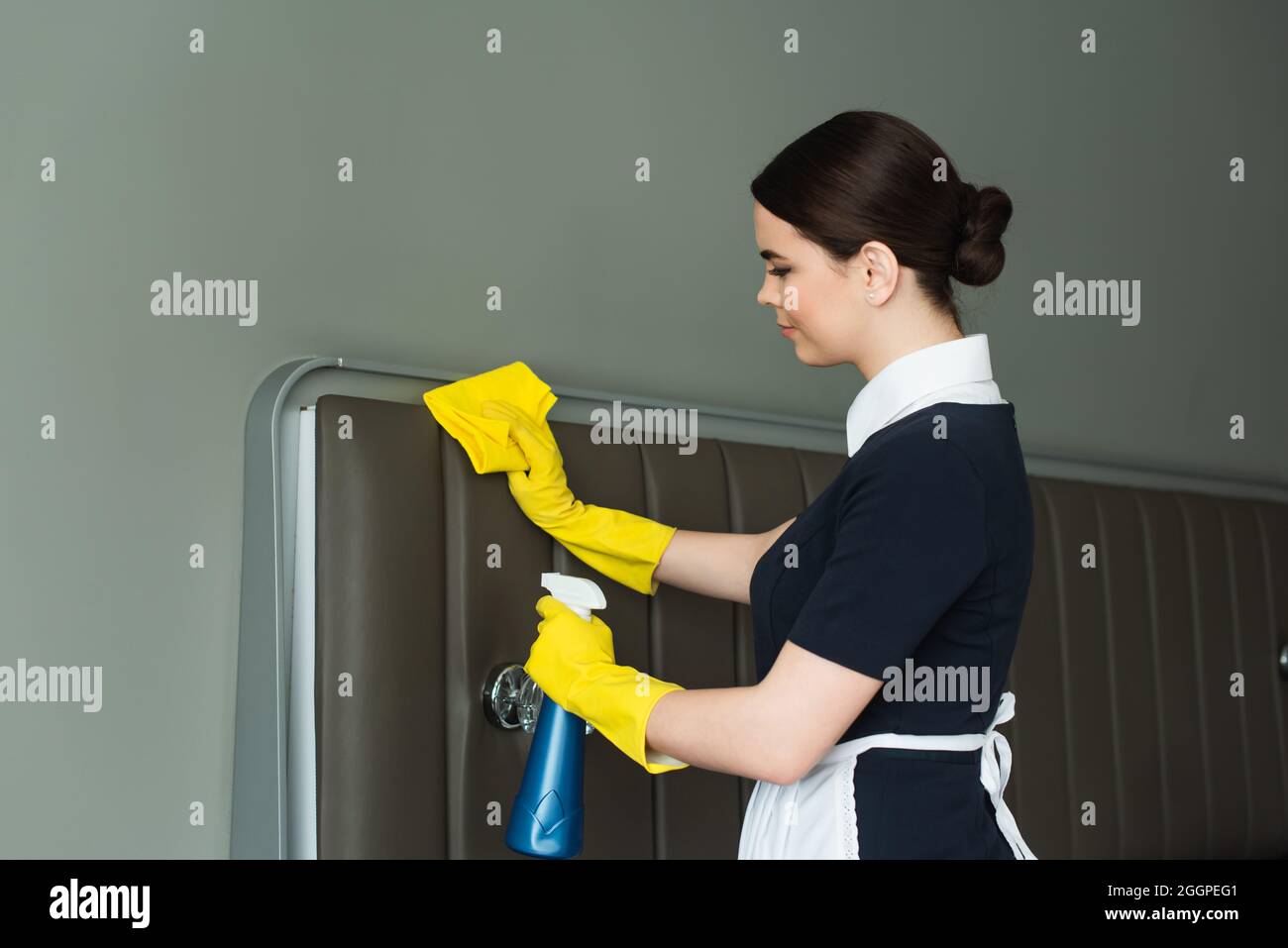 side view of young maid in rubber gloves holding spray bottle and rag ...