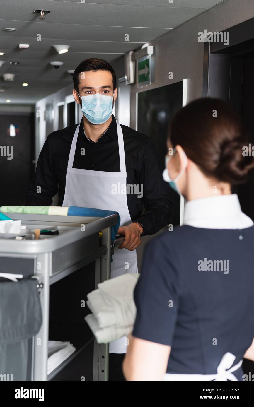 housekeeper in medical mask looking at blurred maid near housekeeping ...