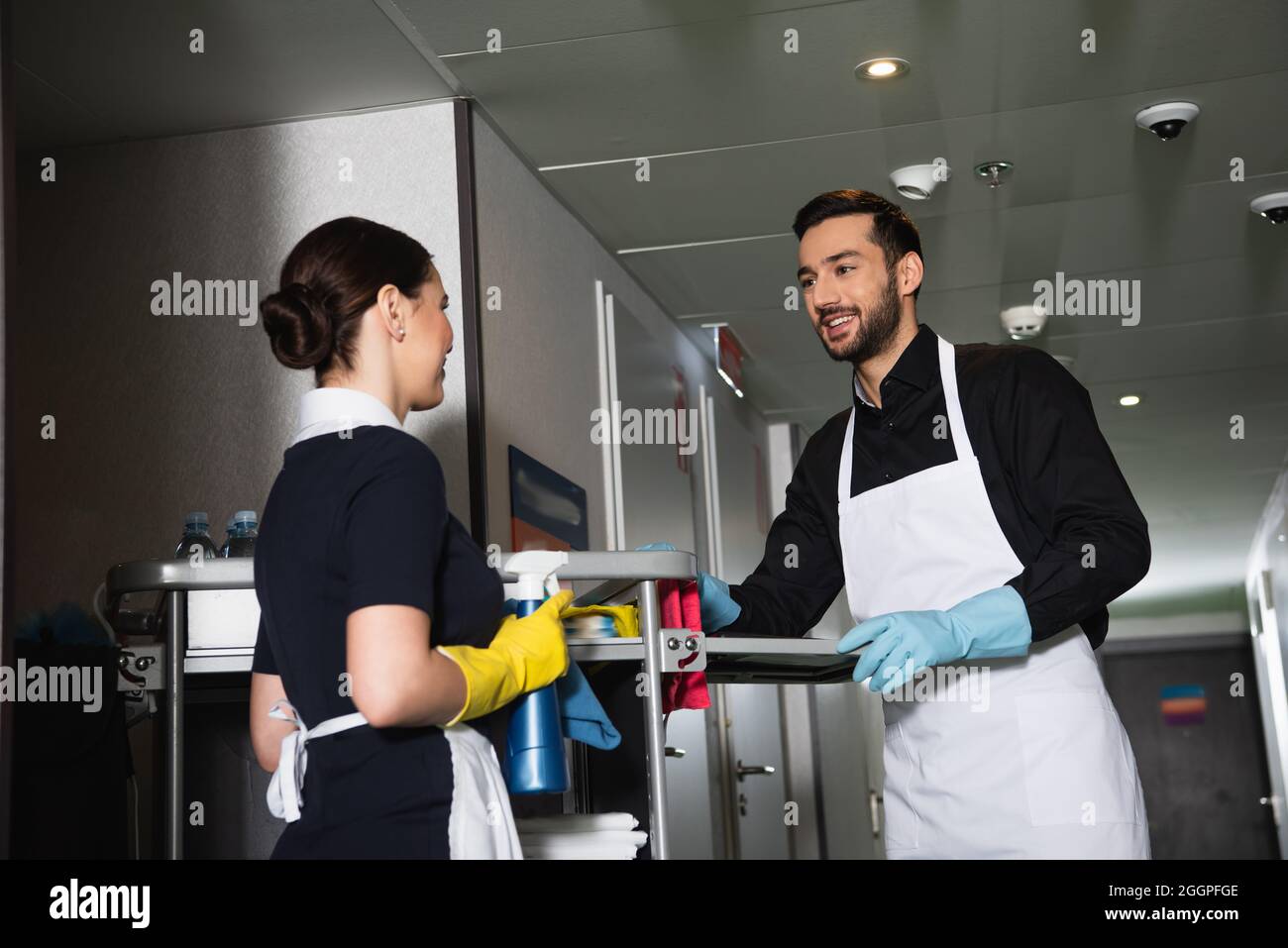 cheerful housekeepers talking near housekeeping cart in corridor of ...