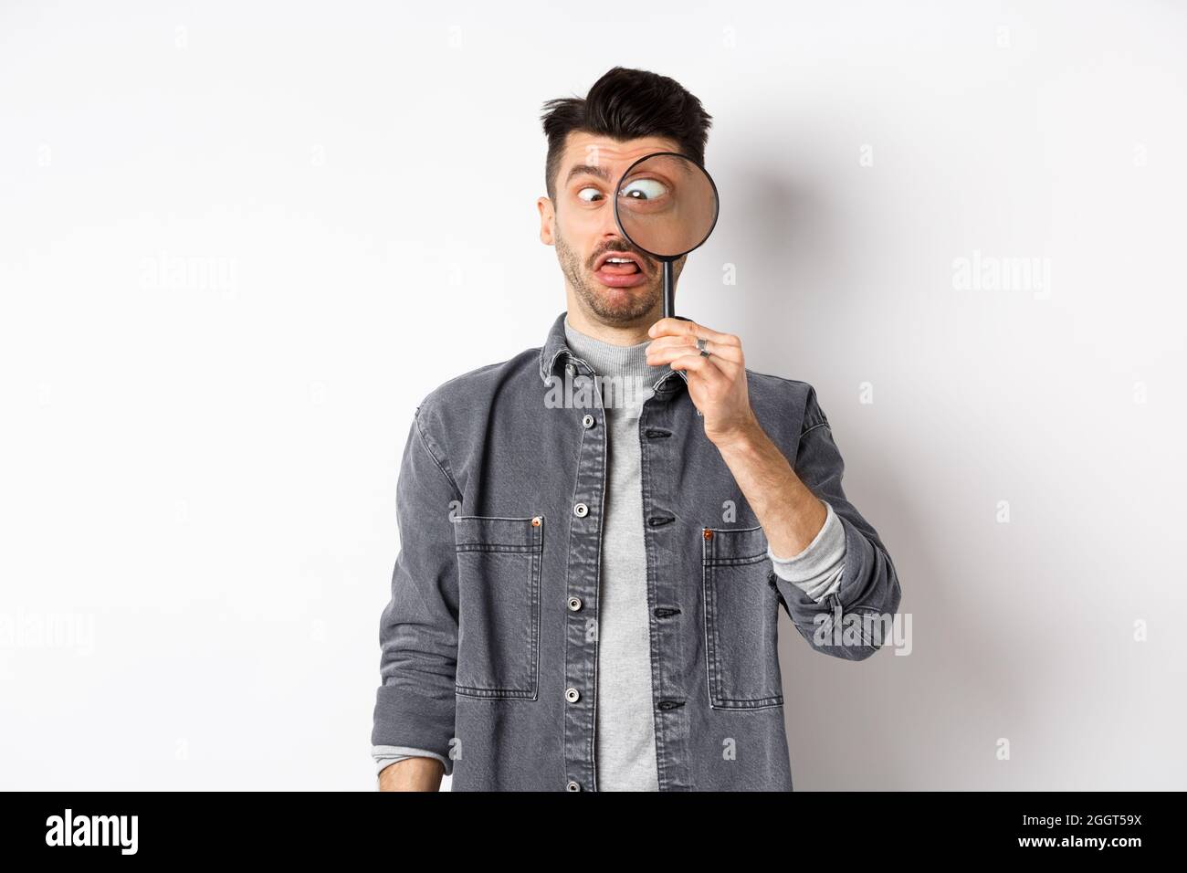 Funny young man squinting, showing faces with magnifying glass ...