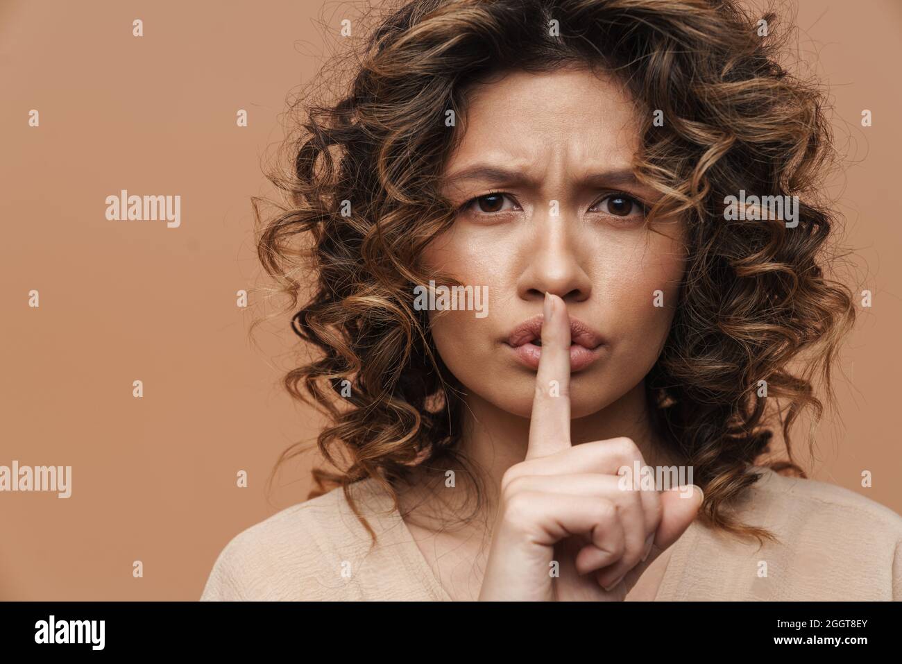 Young hispanic woman frowning while showing silence gesture at camera ...