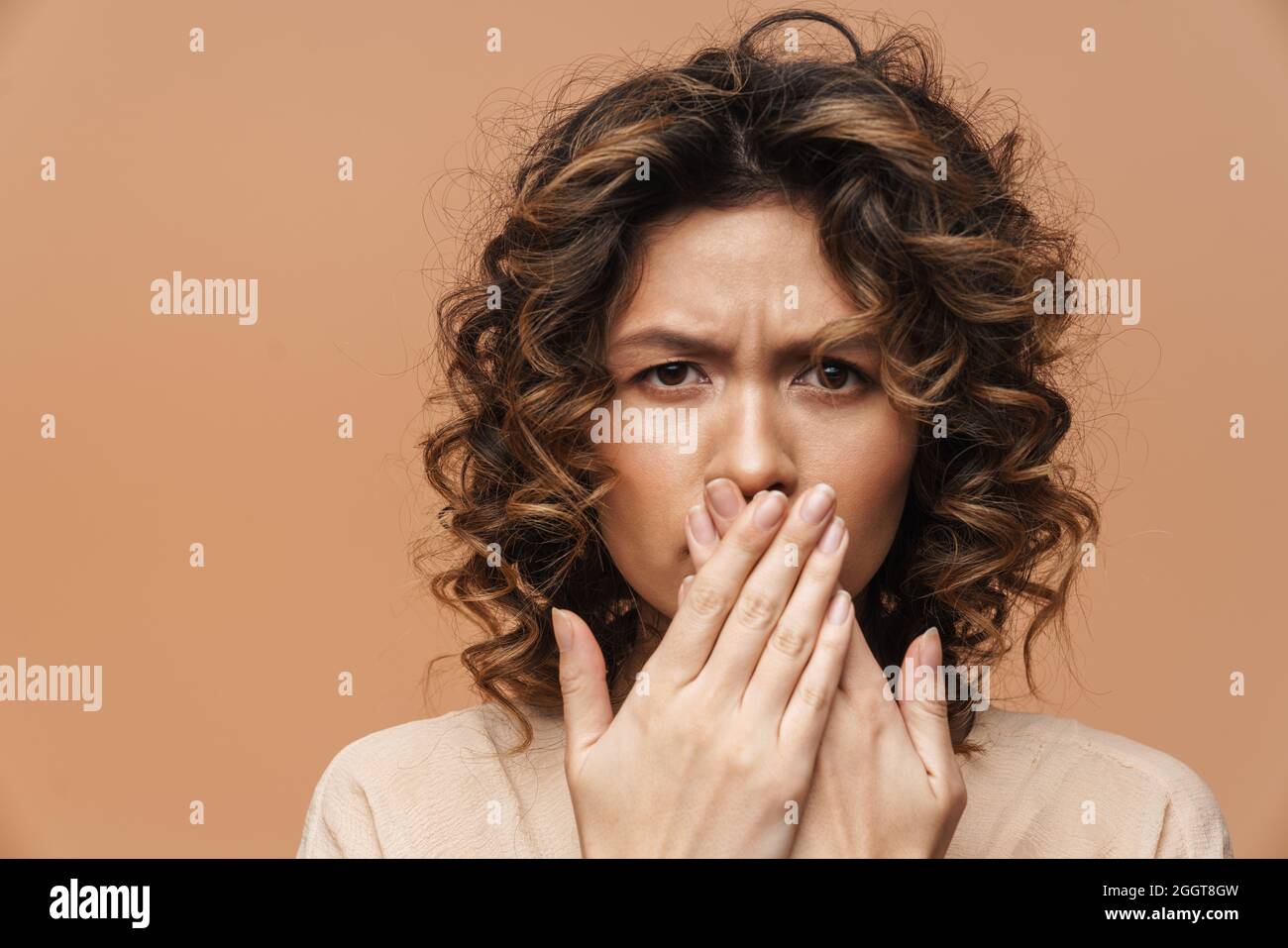 Young curly hispanic woman frowning and covering her mouth isolated ...