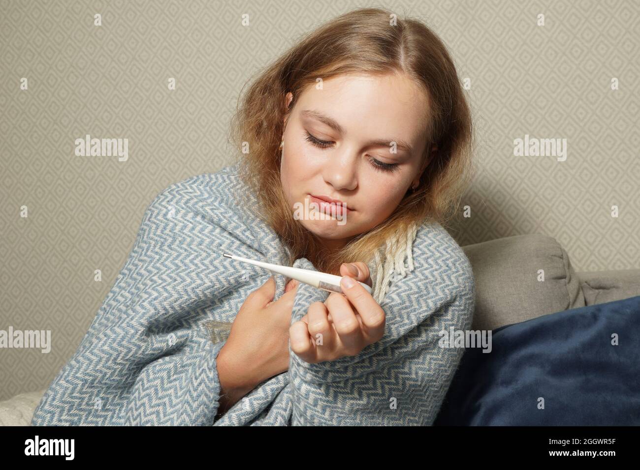 Young blonde woman wrapped in warm coverlet, holds termometer which ...