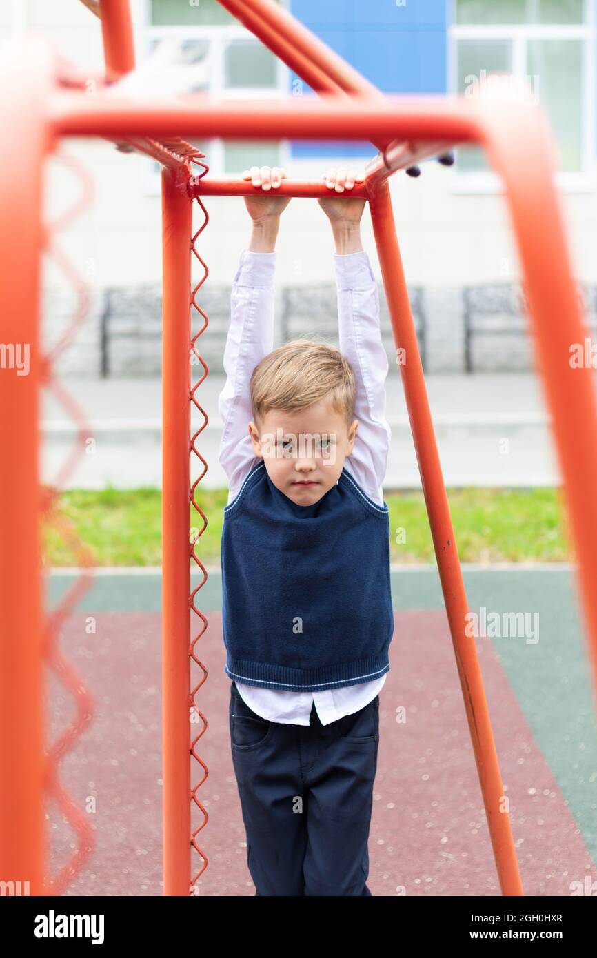 A cute first grader boy at the school on the playground hangs on a ...