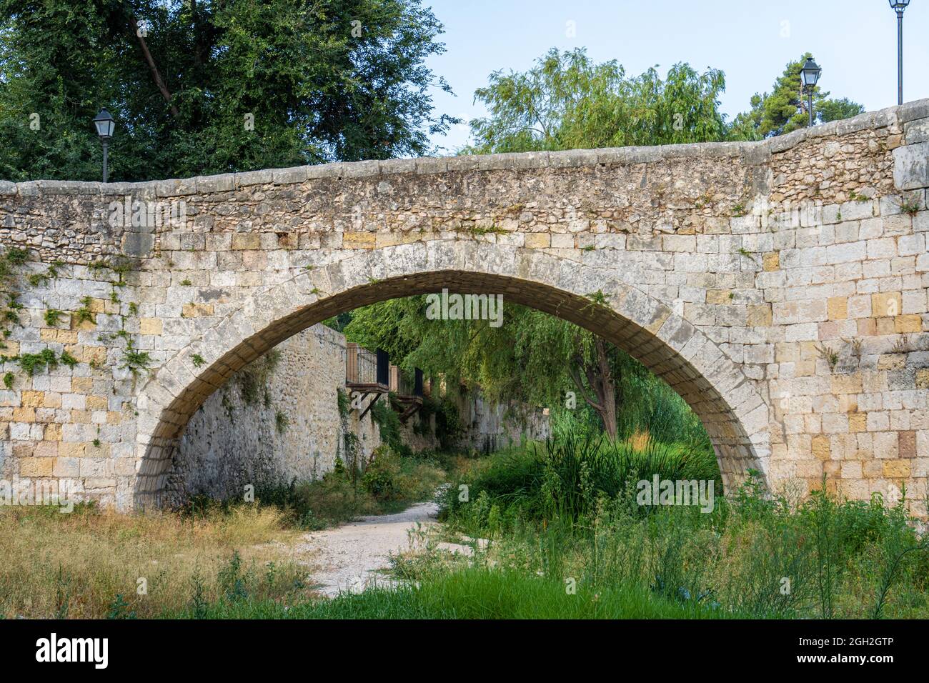 Old stone arched bridge over the walkway with green plants and trees ...