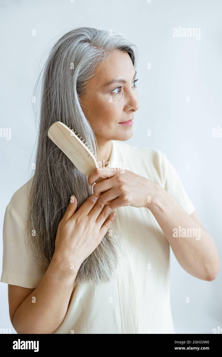 Languid middle aged Asian woman brushes natural hoary hair on light ...