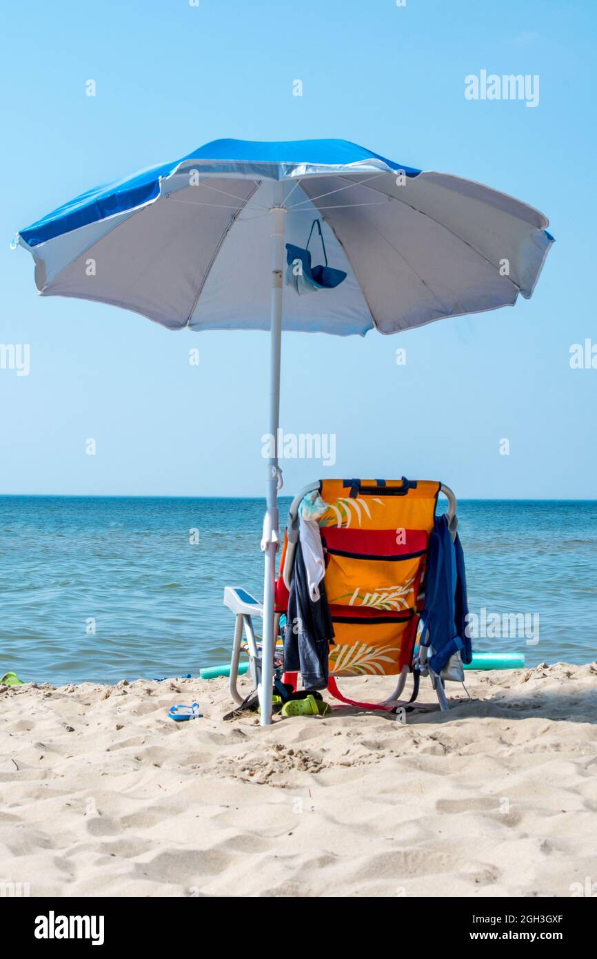 An empty beach chair under a big blue umbrella, is draped with clothing ...