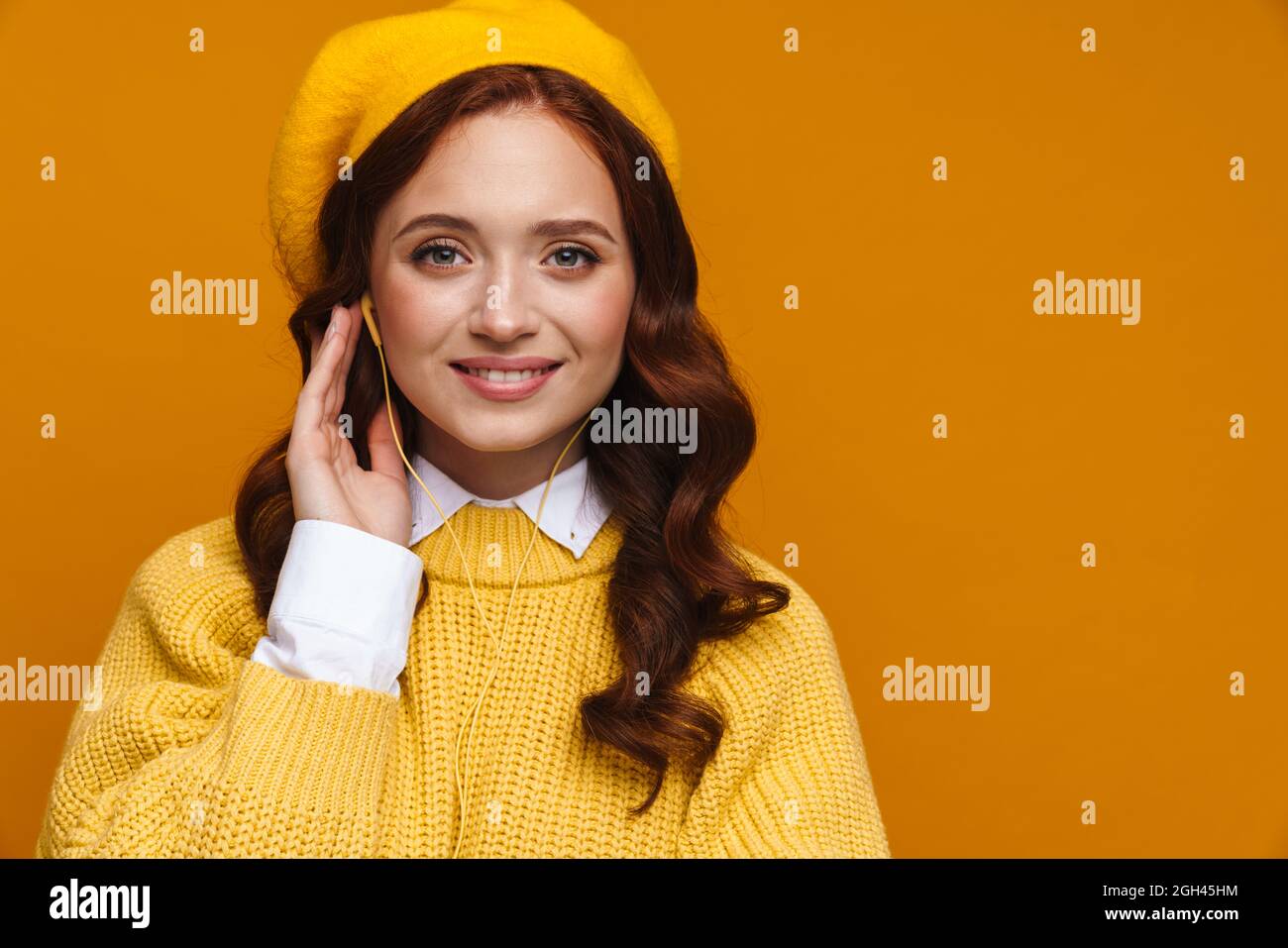 Happy young woman with long red hair in sweater and beret standing over yellow wall background ...