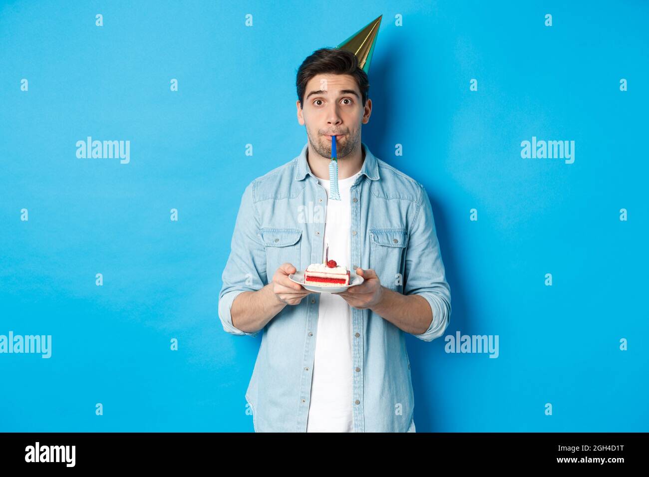 Handsome young man holding birthday cake, wearing party hat and blowing ...
