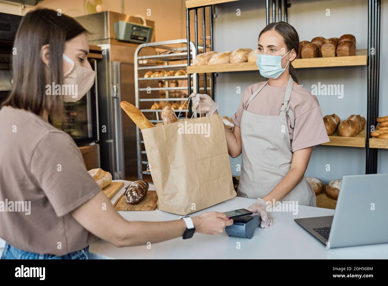 Female baker in mask standing at counter with terminal and giving ...
