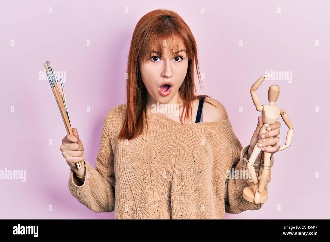 Redhead young woman holding small wooden manikin and painter brushes ...