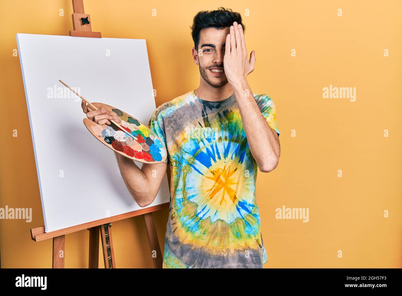 Young hispanic man standing drawing with palette by painter easel stand ...