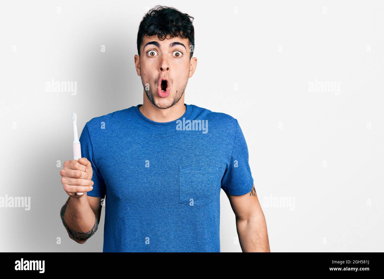 Young hispanic man holding electric toothbrush scared and amazed with ...
